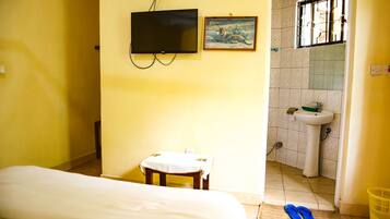 Quarto com cama, uma mesinha, televisão fixada na parede, quadro emoldurado, banheiro com pia e chuveiro, e um par de chinelos no chão.