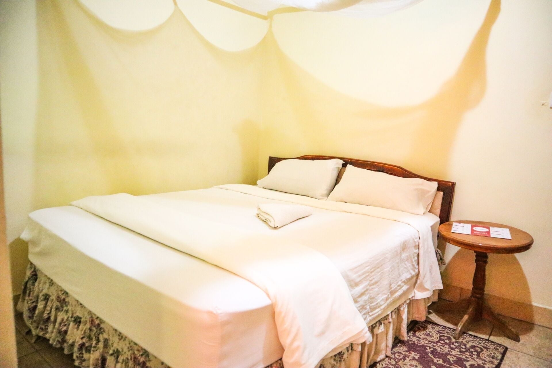 Cama bem arrumada com edredom branco e almofadas, uma pequena mesa de cabeceira com um objeto vermelho em cima, e um tapete estampado no chão.