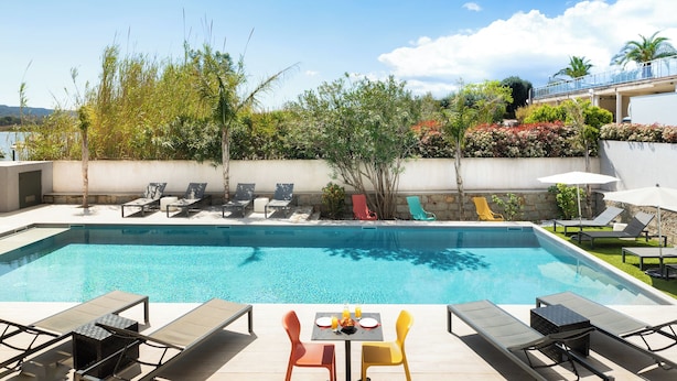 Piscina retangular com espreguiçadeiras e uma mesa, rodeada por vegetação e céu claro.