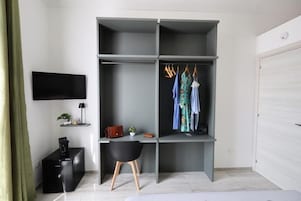 Quarto moderno com guarda-roupa cinzento, escrivaninha, cadeira preta, televisão e porta branca.