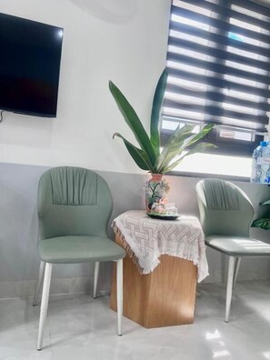 Quarto com duas cadeiras, uma pequena mesa de madeira com um objeto decorativo e uma televisão fixada na parede.
