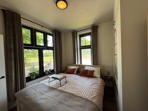 Quarto com cama, mesa de cabeceira, janela com cortinas e vista para o verde lá fora.