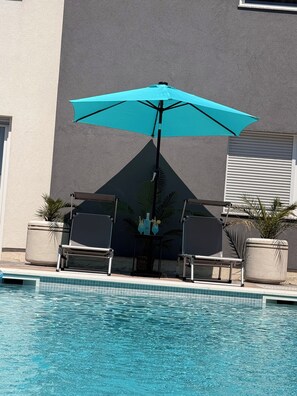 Uma área de piscina com um guarda-sol turquesa, duas espreguiçadeiras e plantas em vasos.