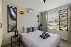 Quarto com cama, duas luminárias na parede, um vaso de planta e janelas amplas com persianas.