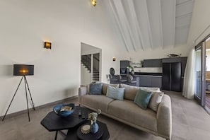Uma sala de estar moderna com sofá, mesa de centro e abajur. É visível uma área de cozinha através de uma porta aberta.