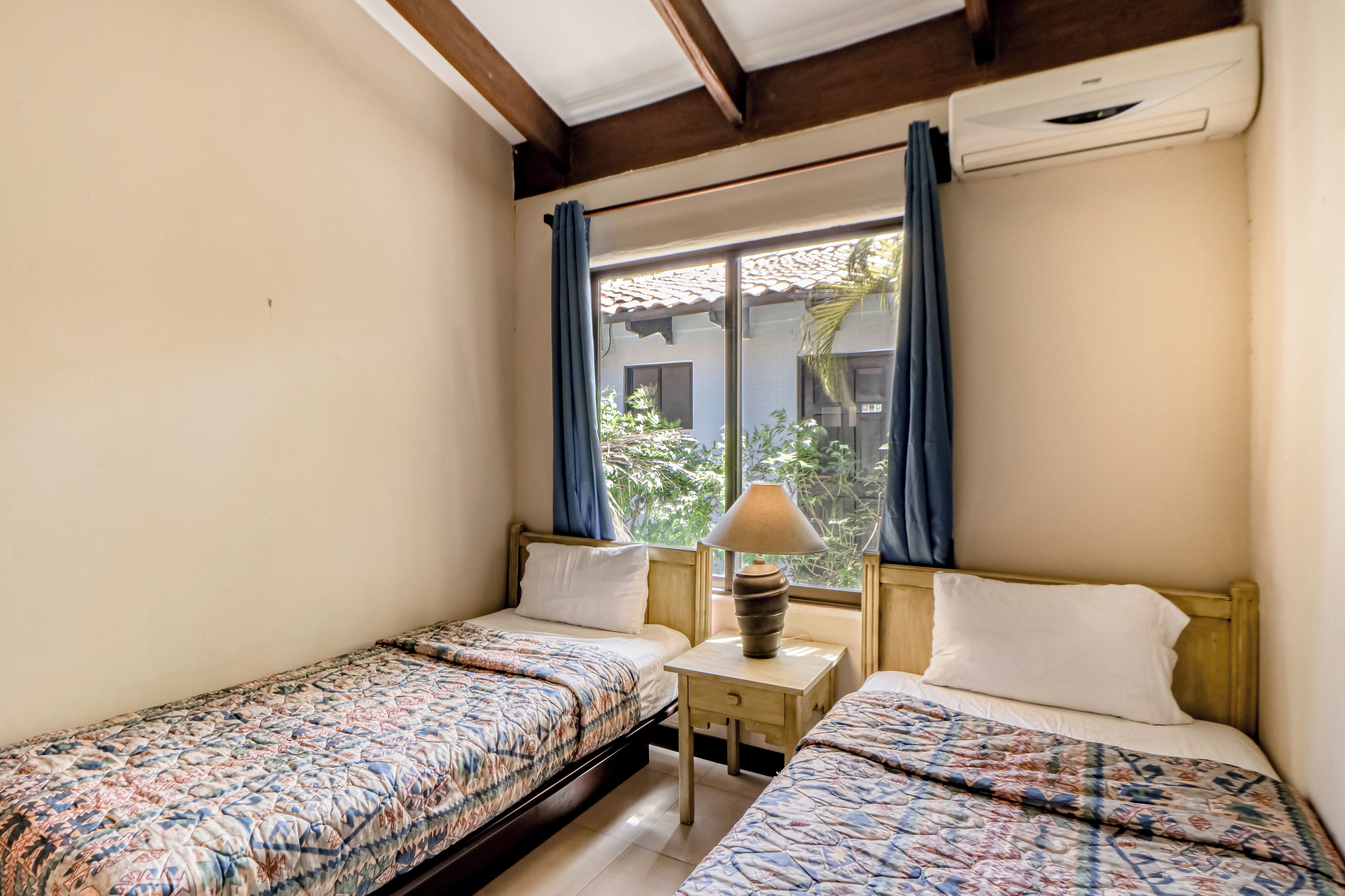 Quarto com duas camas de solteiro, uma mesinha com abajur e uma janela com cortinas.
