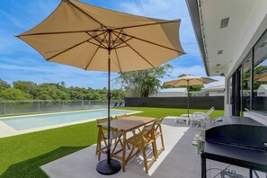 Um quintal com piscina, mesa de madeira com cadeiras e churrasqueira.