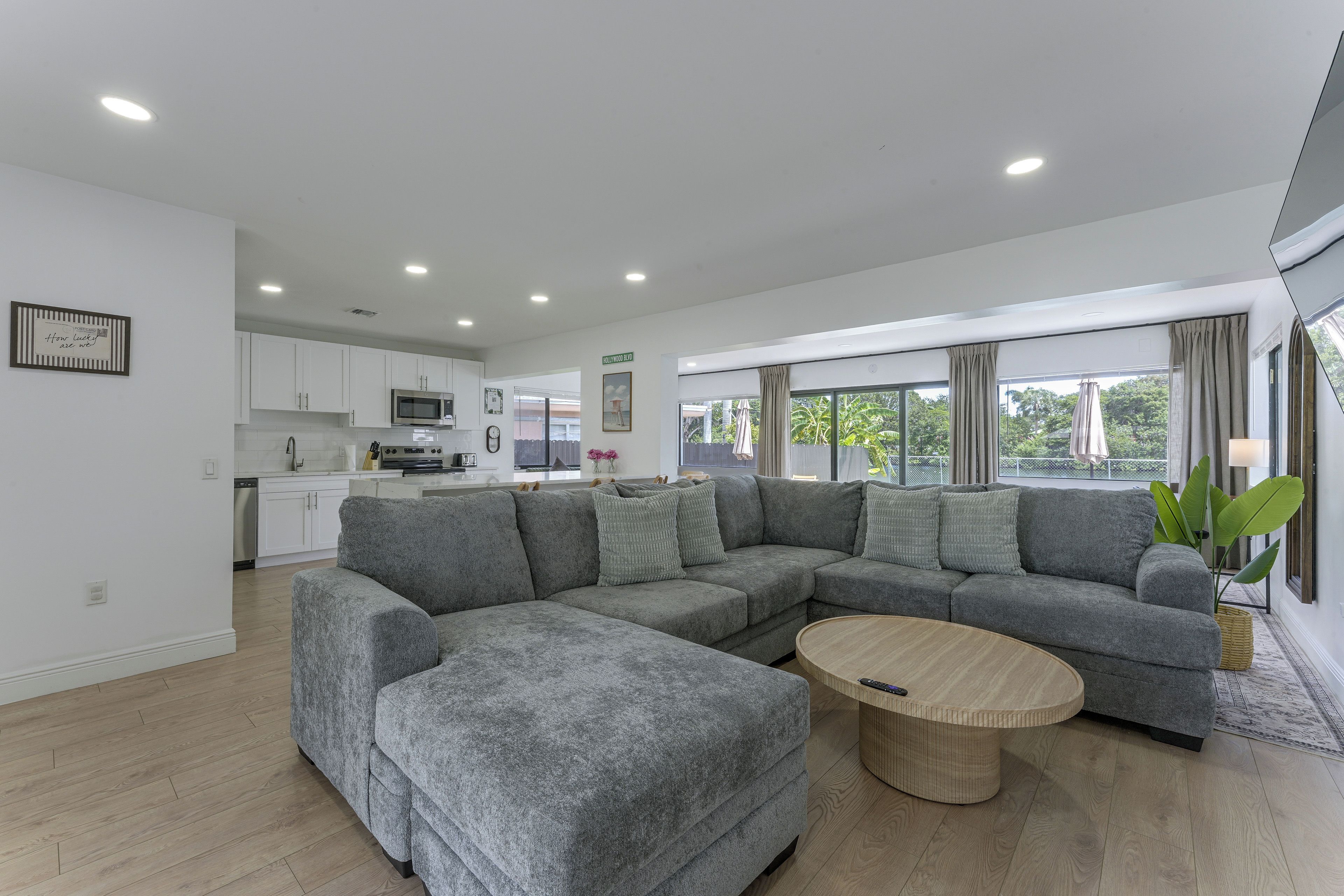 Uma sala de estar moderna com um sofá seccional cinza, uma mesa de centro de madeira redonda e vista para a cozinha ao fundo.