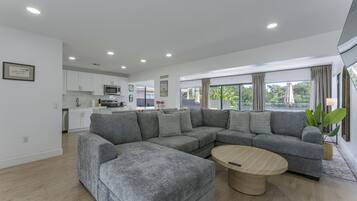 Uma sala de estar moderna com um sofá seccional cinza, uma mesa de centro de madeira redonda e vista para a cozinha ao fundo.