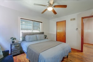 Quarto com cama, mesa de cabeceira, ventoinha no teto, janela com persianas e porta de madeira.