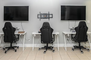 Quarto com três cadeiras de jogos, duas mesas e duas grandes televisões de tela plana fixadas na parede.