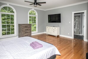Quarto com cama grande, penteadeira, televisão e duas janelas.