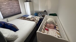 Quarto com cama, berço, radiador, janela com persianas, guarda-roupa e uma pequena mesa com cadeira.