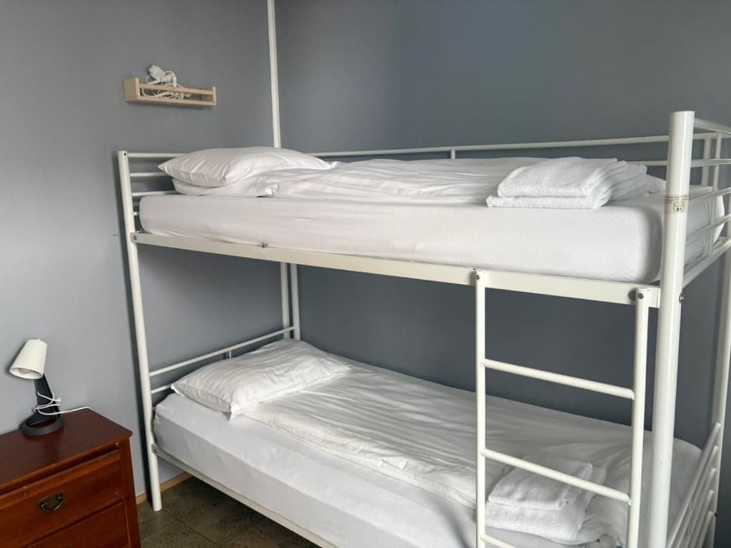 Uma cama de beliche com roupa de cama branca, uma mesa de cabeceira com abajur e uma pequena prateleira com um objeto decorativo.