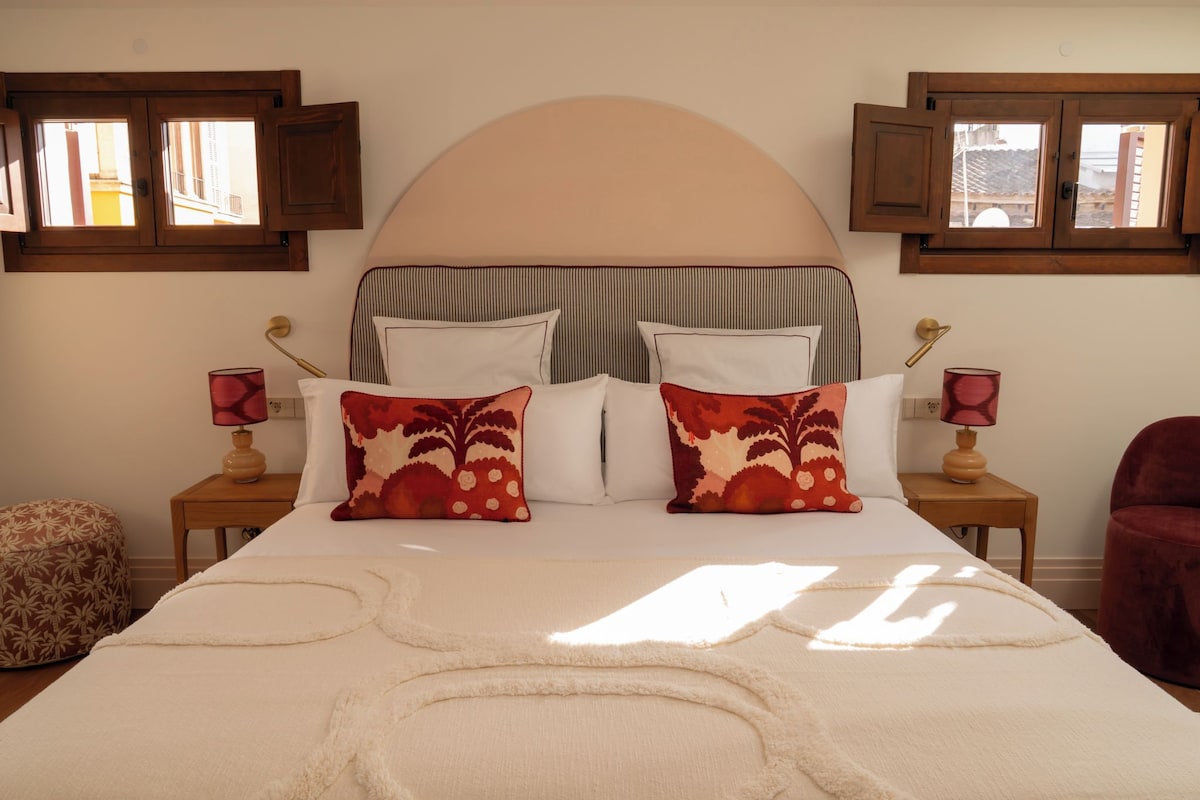 Uma cama com cabeceira, duas mesas de cabeceira com abajures e uma janela com cortinas.