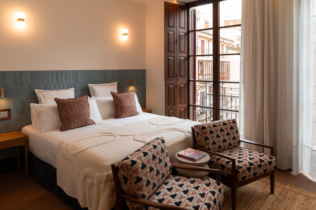 Quarto de hotel com cama, duas mesas de cabeceira, uma cadeira, uma mesinha e janela com vista para edifícios.