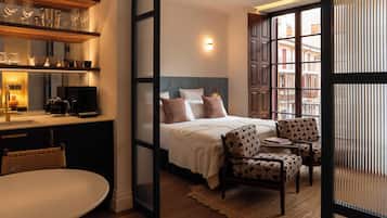 Quarto de hotel com cama, área de estar com cadeiras, máquina de café e prateleira com copos e xícaras.