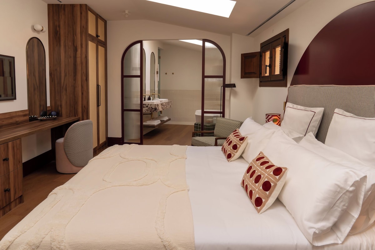 Quarto com cama grande, escrivaninha, cadeira e espelho. Vê-se um banheiro e um corredor.