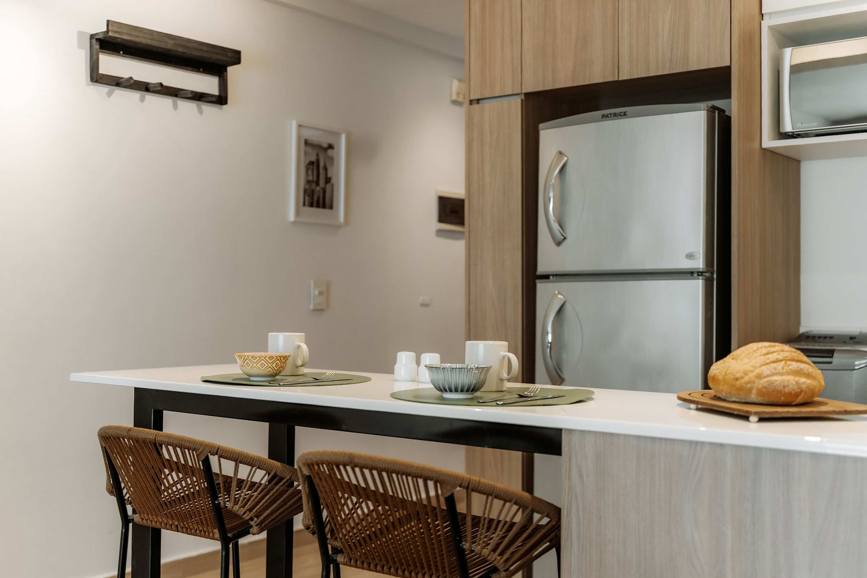 Uma cozinha moderna com uma geladeira de portas duplas, duas cadeiras de vime, uma bancada branca e uma barra de pão sobre uma tábua de corte.