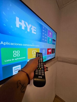 Mão segurando um controle remoto em frente a uma televisão exibindo uma interface de usuário com vários ícones de aplicativos.