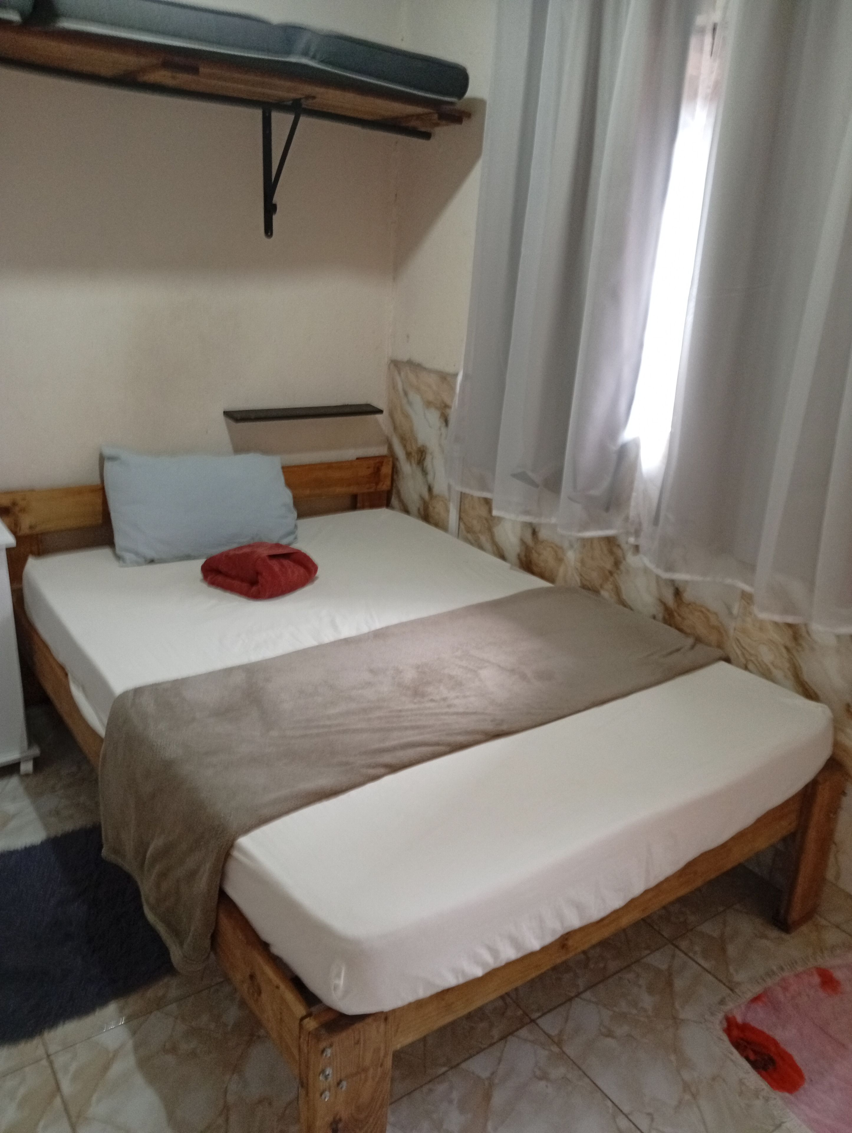 Cama de solteiro com estrutura de madeira, colchão branco e manta vermelha. Há uma prateleira de madeira acima da cama e uma janela com cortinas.