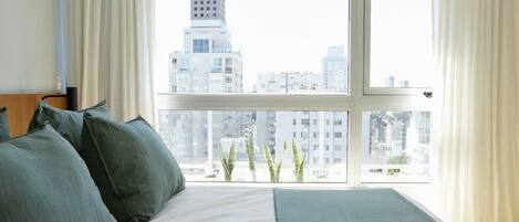 Quarto de hotel com cama, cortinas e vista para edifícios da cidade.