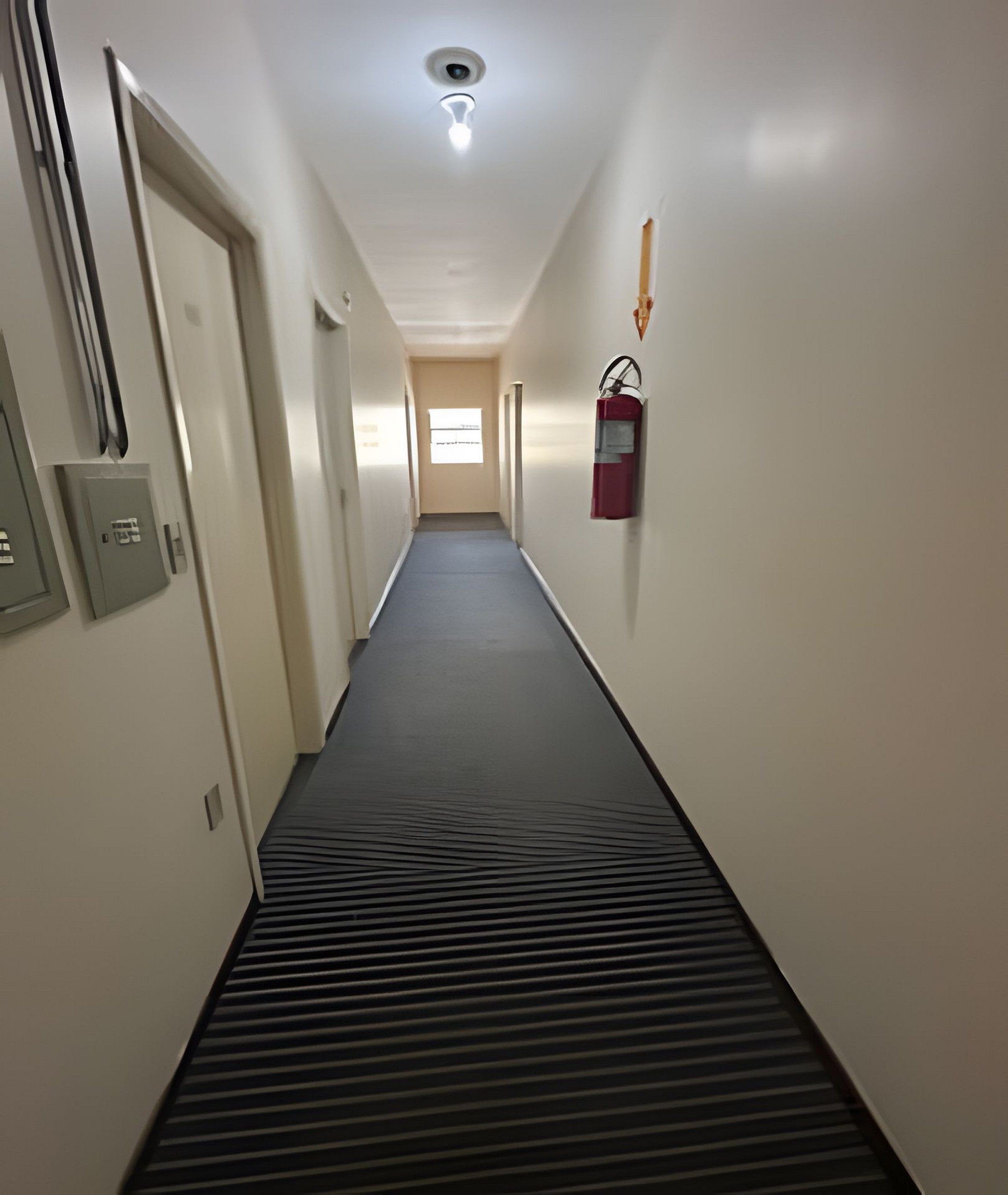 Corredor estreito com um extintor preso na parede, uma saída de ventilação no chão e uma porta à esquerda.