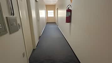 Corredor estreito com um extintor preso na parede, uma saída de ventilação no chão e uma porta à esquerda.