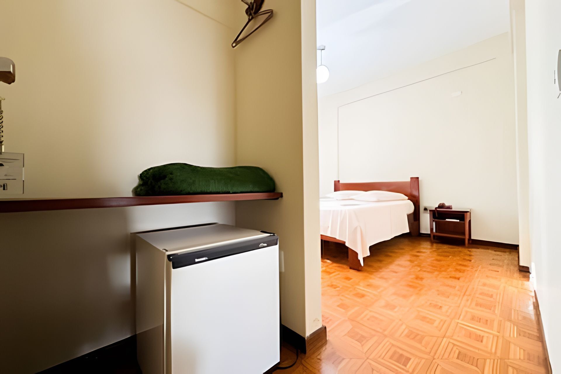 Quarto compacto com cama, escrivaninha pequena, geladeira e prateleira com um objeto verde.