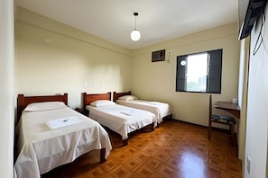 Quarto com três camas de solteiro, piso de madeira, janela com persianas e uma pequena escrivaninha.