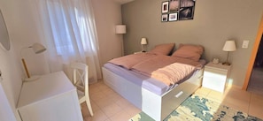 Um quarto com cama, mesas de cabeceira, uma cadeira e janela com cortinas.