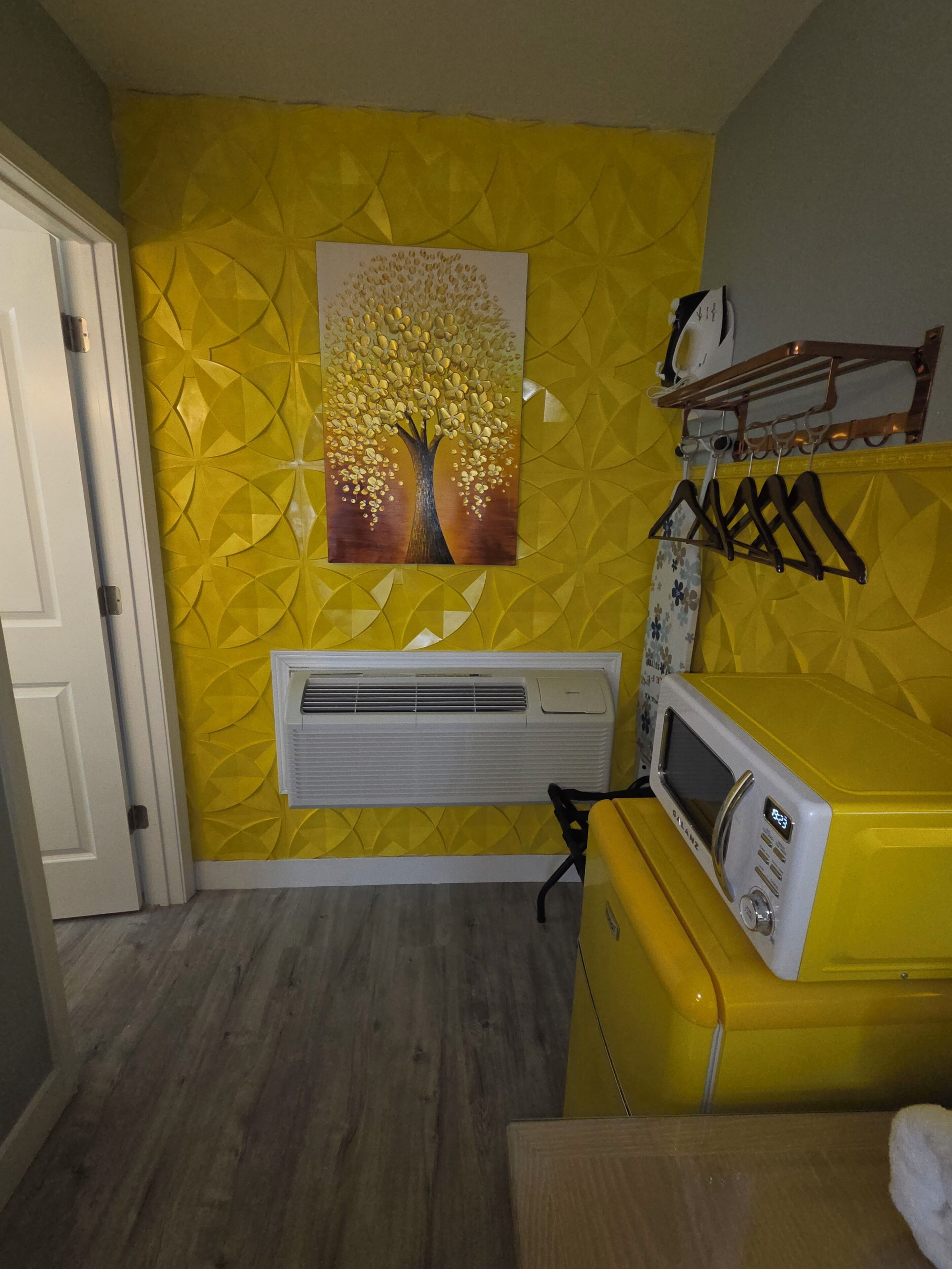 Quarto com design geométrico amarelo na parede, ar condicionado branco, micro-ondas e um suporte de madeira para cabides.