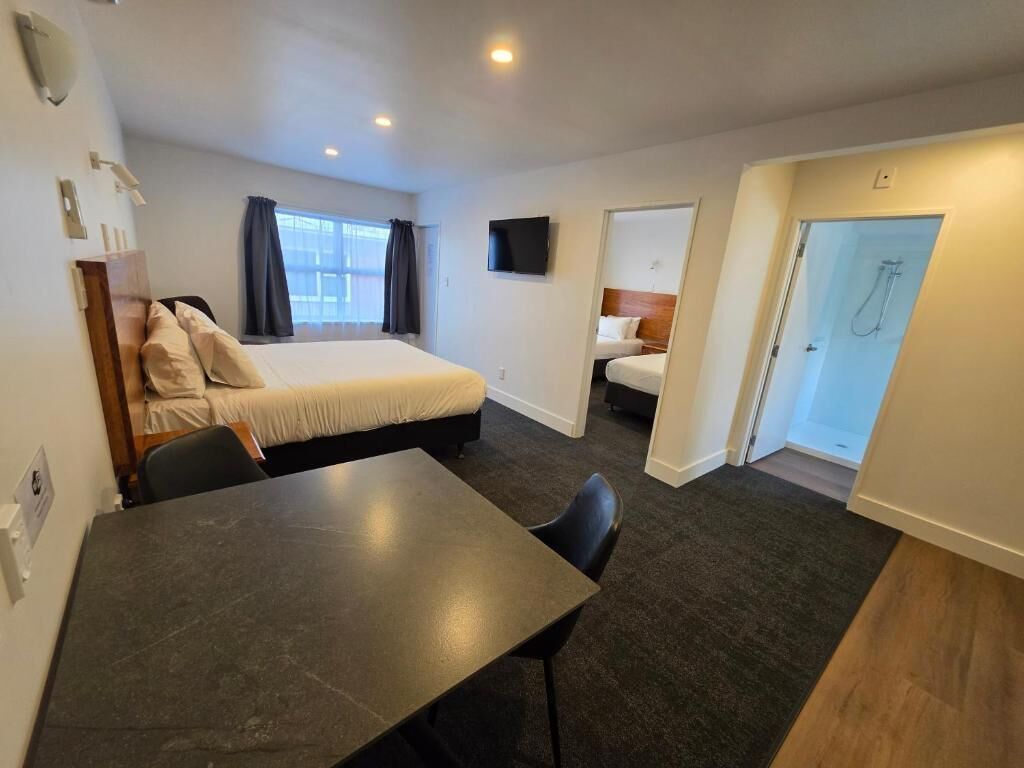 Quarto de hotel com cama, escrivaninha, cadeira, televisão e chuveiro.