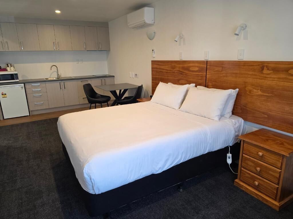 Quarto de hotel com cama, mesa de cabeceira e área de cozinha compacta.