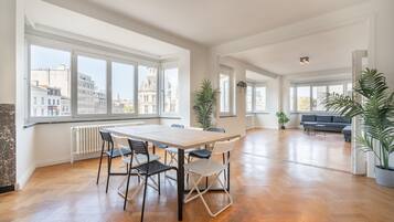 Ampla sala de estar com grande mesa de jantar, várias cadeiras e um sofá. Há algumas plantas em vasos e vista para edifícios por grandes janelas.