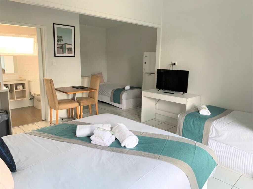 Quarto de hotel com duas camas, uma escrivaninha, televisão e banheiro visível através de uma porta aberta.