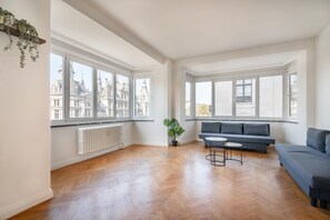 Uma sala de estar ampla com grandes janelas, um radiador, um sofá cinzento e uma mesa pequena.