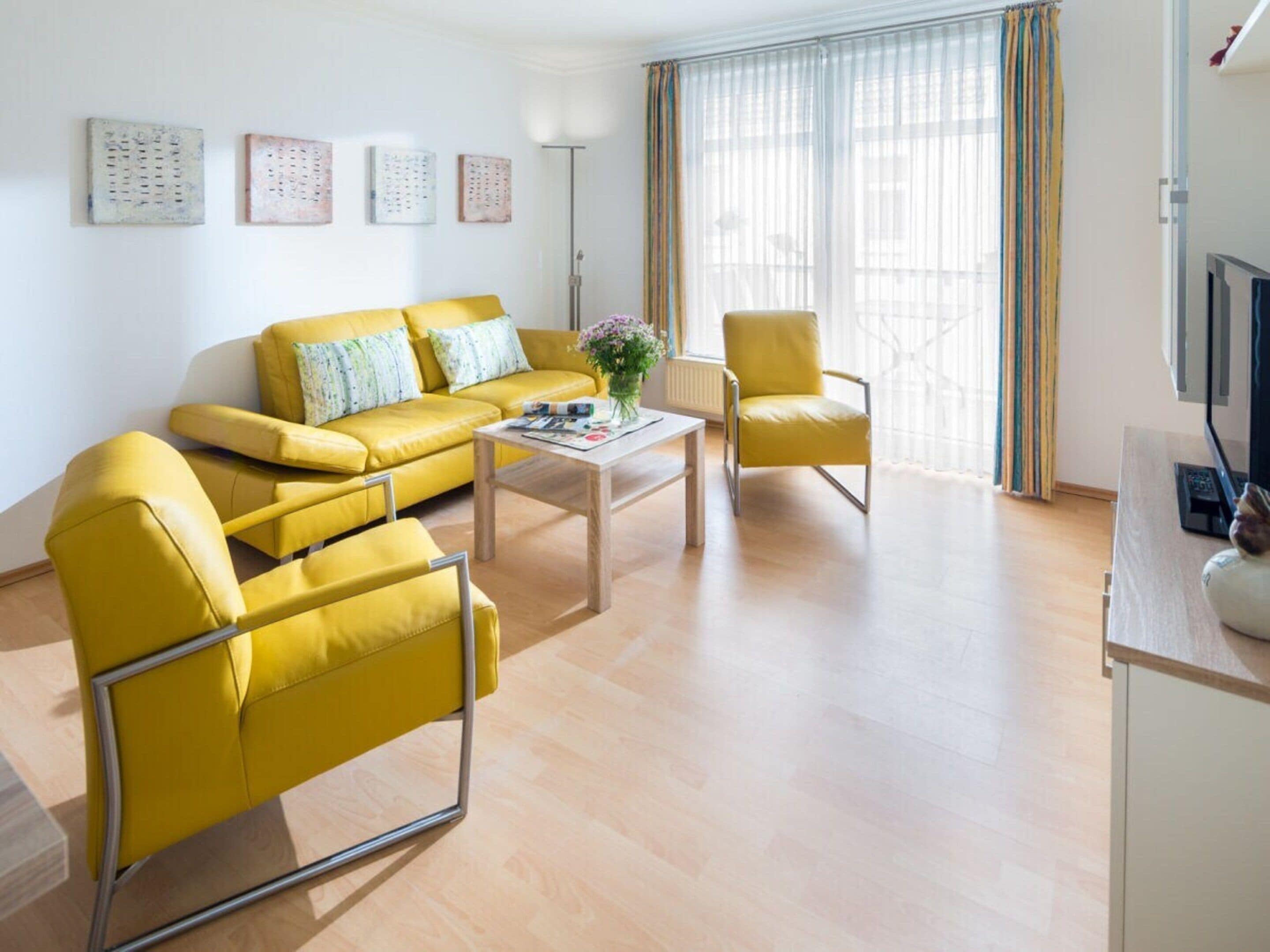 Uma sala de estar moderna com um sofá e uma poltrona amarelos, e uma mesa de centro. Há quatro obras de arte penduradas na parede e um abajur.