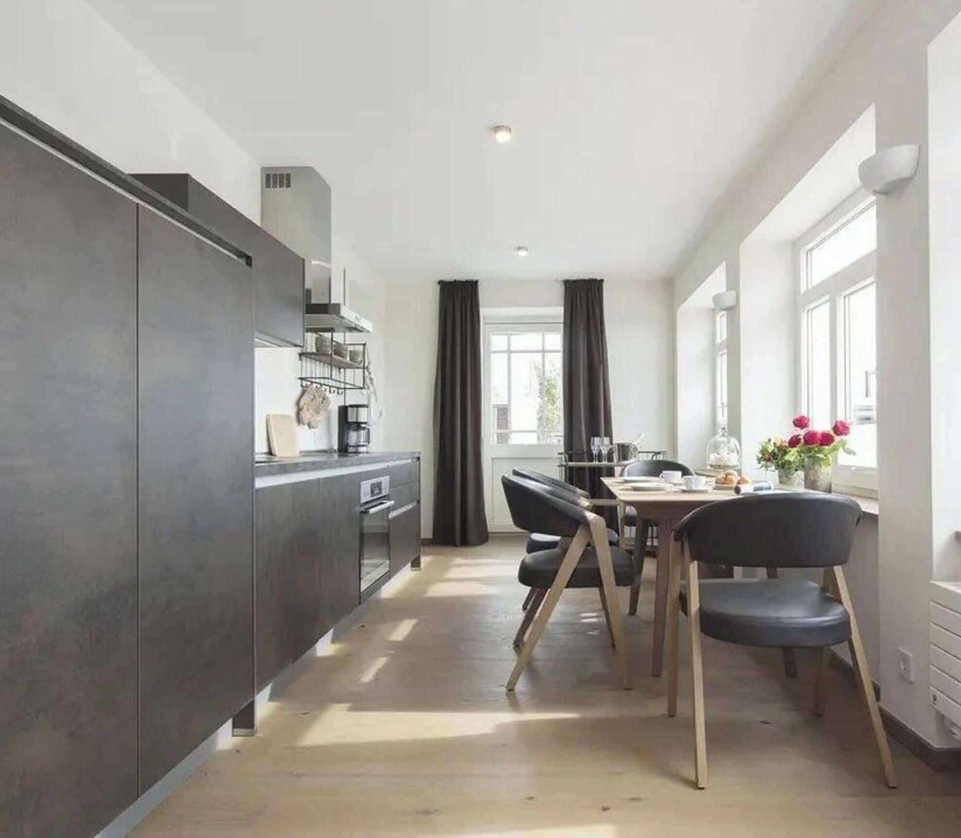 Cozinha moderna com armários escuros, área de jantar com mesa e cadeiras, e janela com cortinas.