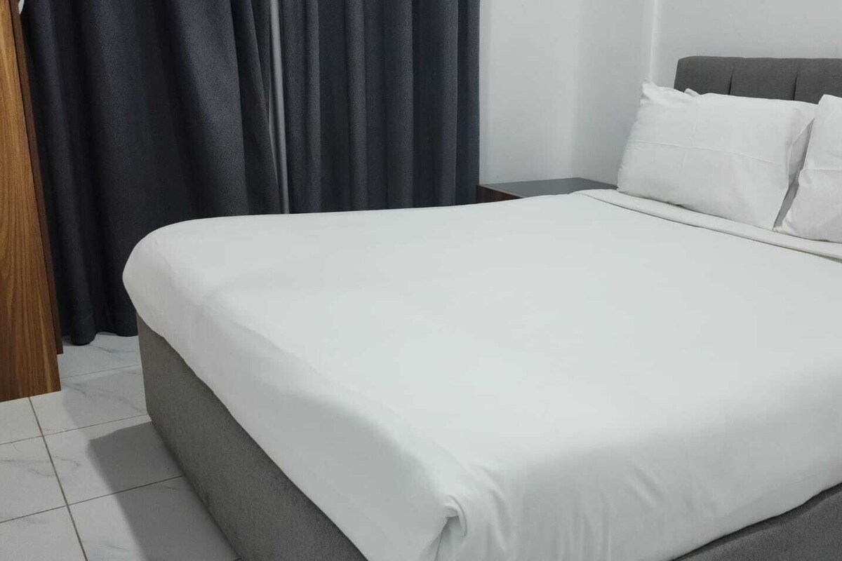 Cama com cabeceira cinza e roupa de cama branca num quarto com porta de madeira e cortinas escuras.