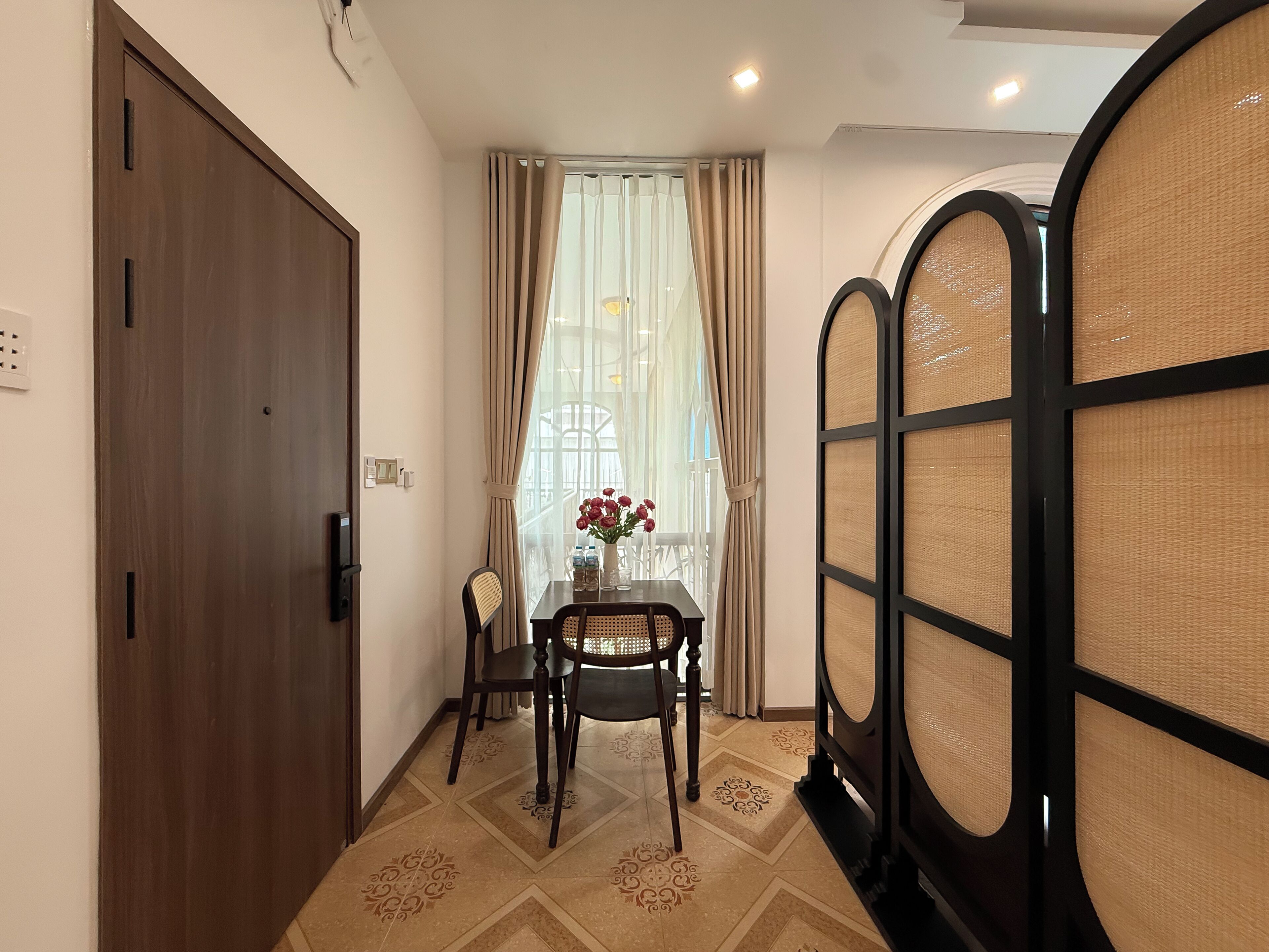 Um quarto com porta de madeira, uma mesa com cadeiras, janela com cortinas e um painel decorativo na parede.