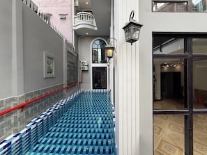 Um corredor interno estreito com chão de azulejos quadriculado em azul e branco, corrimão vermelho e um portão preto no final.