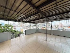 Varanda ampla com cobertura metálica e piso de azulejos, com vista para uma paisagem urbana com edifícios e áreas verdes.