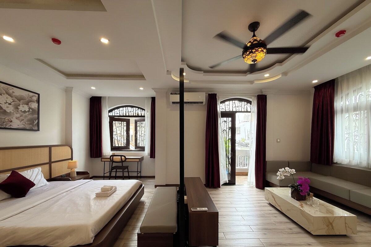 Quarto de hotel com cama, escrivaninha, banco e vista pela janela com cortinas.