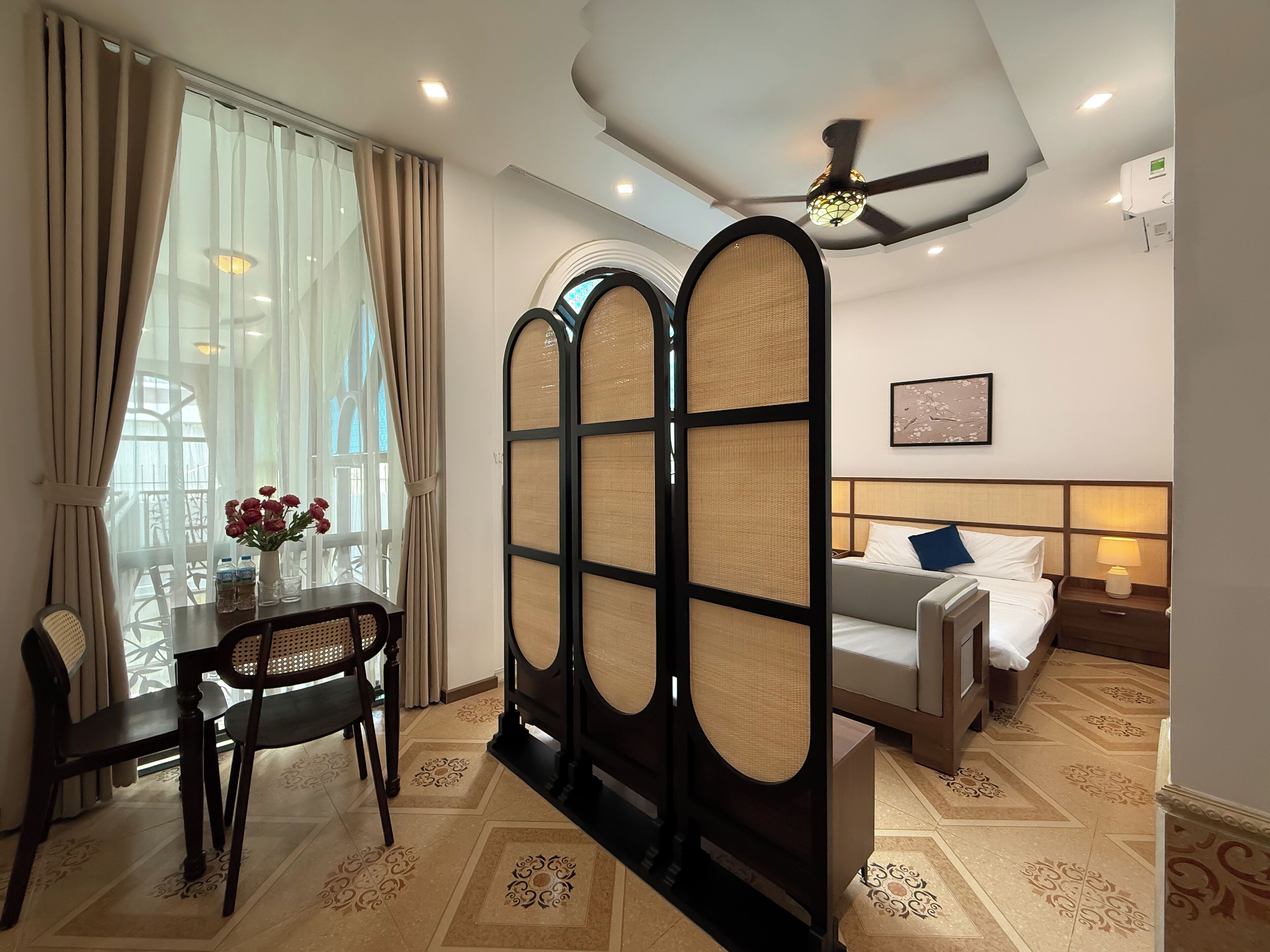 Quarto de hotel com cama, escrivaninha com cadeira, biombos decorativos, ventoinha no teto e vista pela janela.