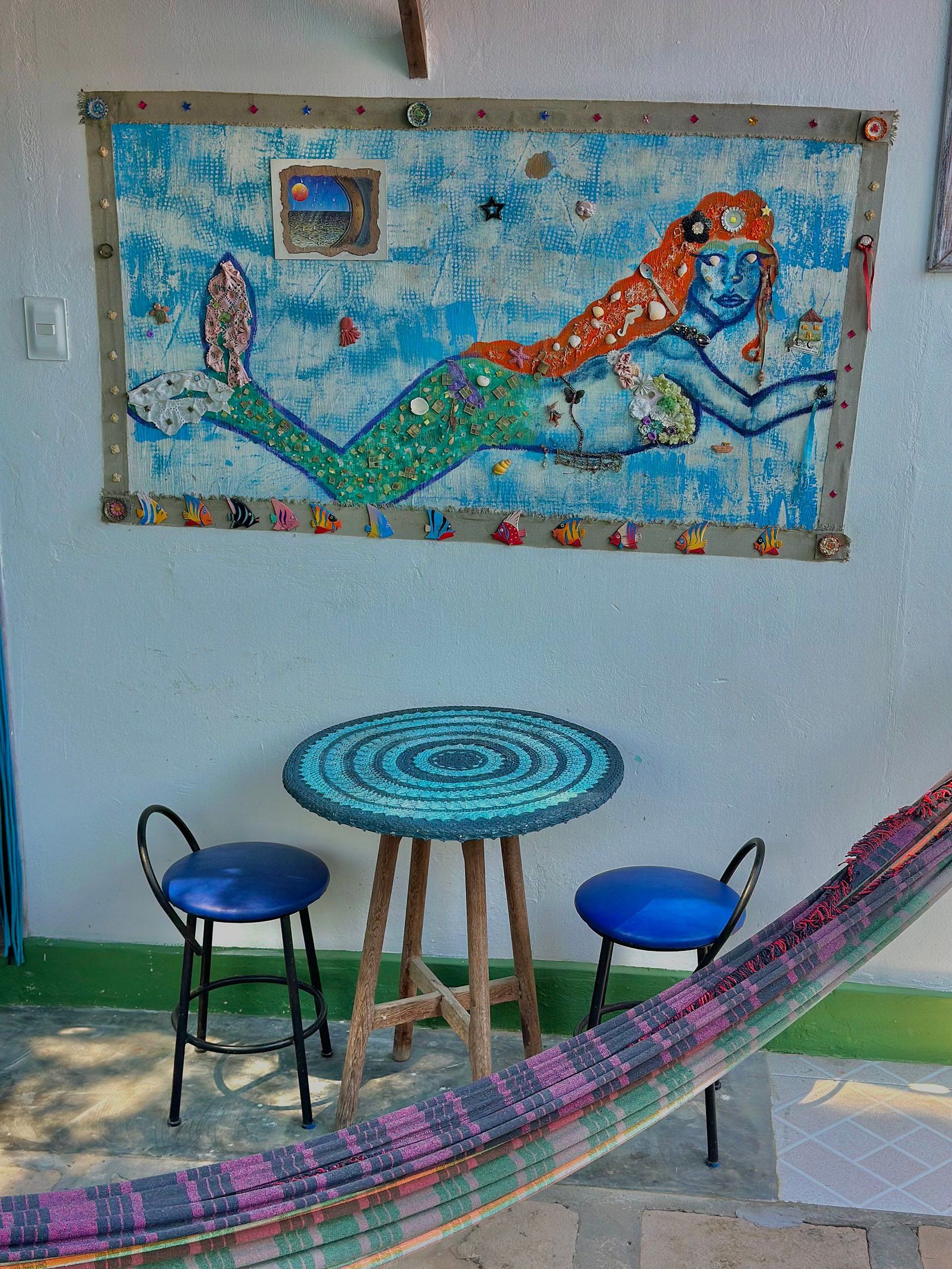 Uma rede, duas cadeiras azuis e uma mesa redonda com uma obra de mosaico na parede.