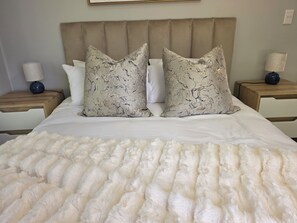 Uma cama com edredom branco e duas almofadas decorativas, ladeada por mesas de cabeceira de madeira com luminárias.