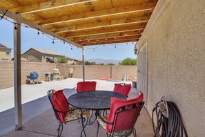 Um pátio externo com mesa e cadeiras redondas, uma pérgola acima e vista para uma área residencial com casas e montanhas ao fundo.