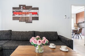 Uma sala de estar com um sofá cinza escuro, uma mesa de centro de madeira, um vaso com rosas cor-de-rosa, e uma xícara e pires florais.
