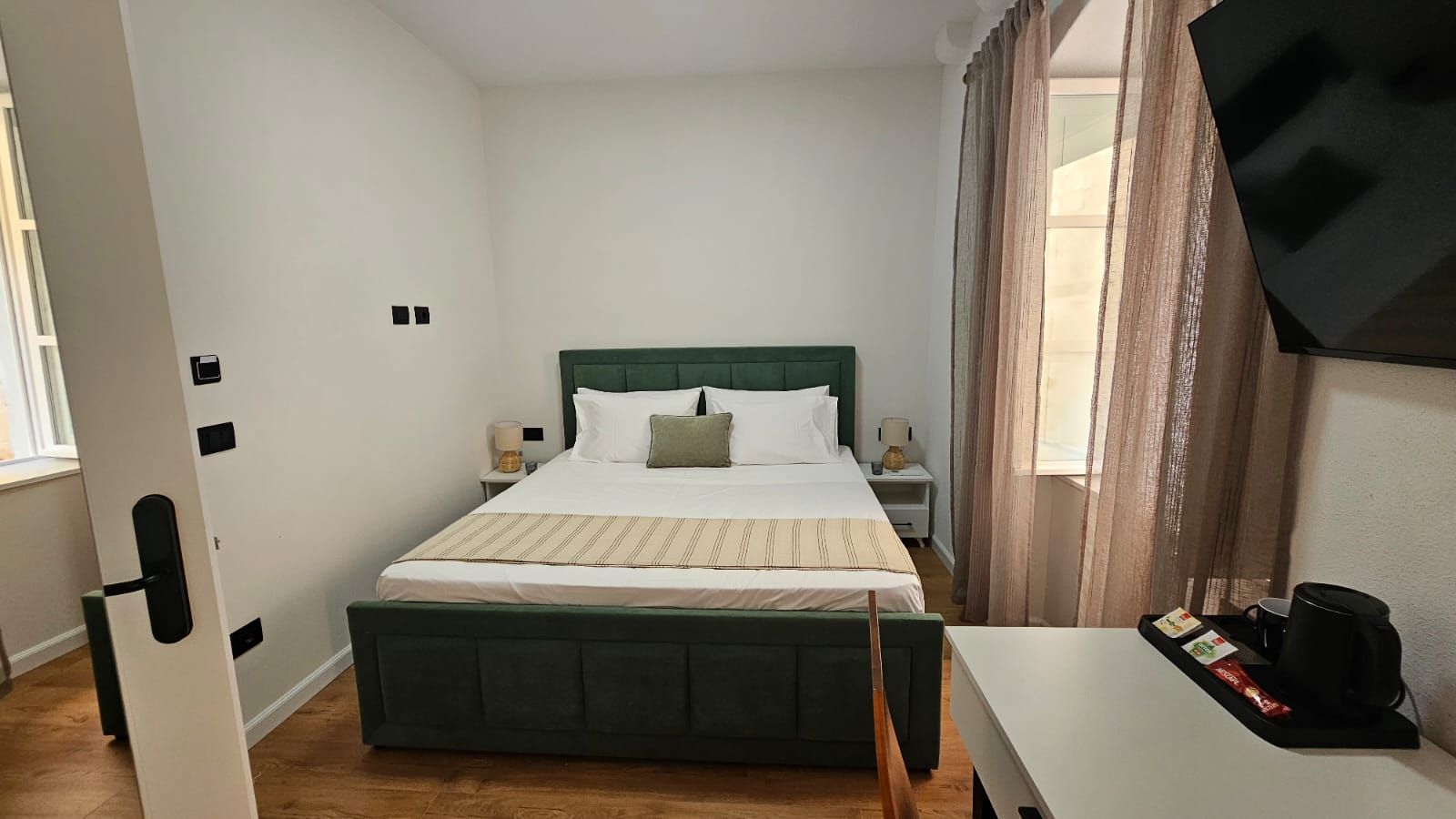 Quarto de hotel com cama, mesas de cabeceira, escrivaninha e janela com cortinas.